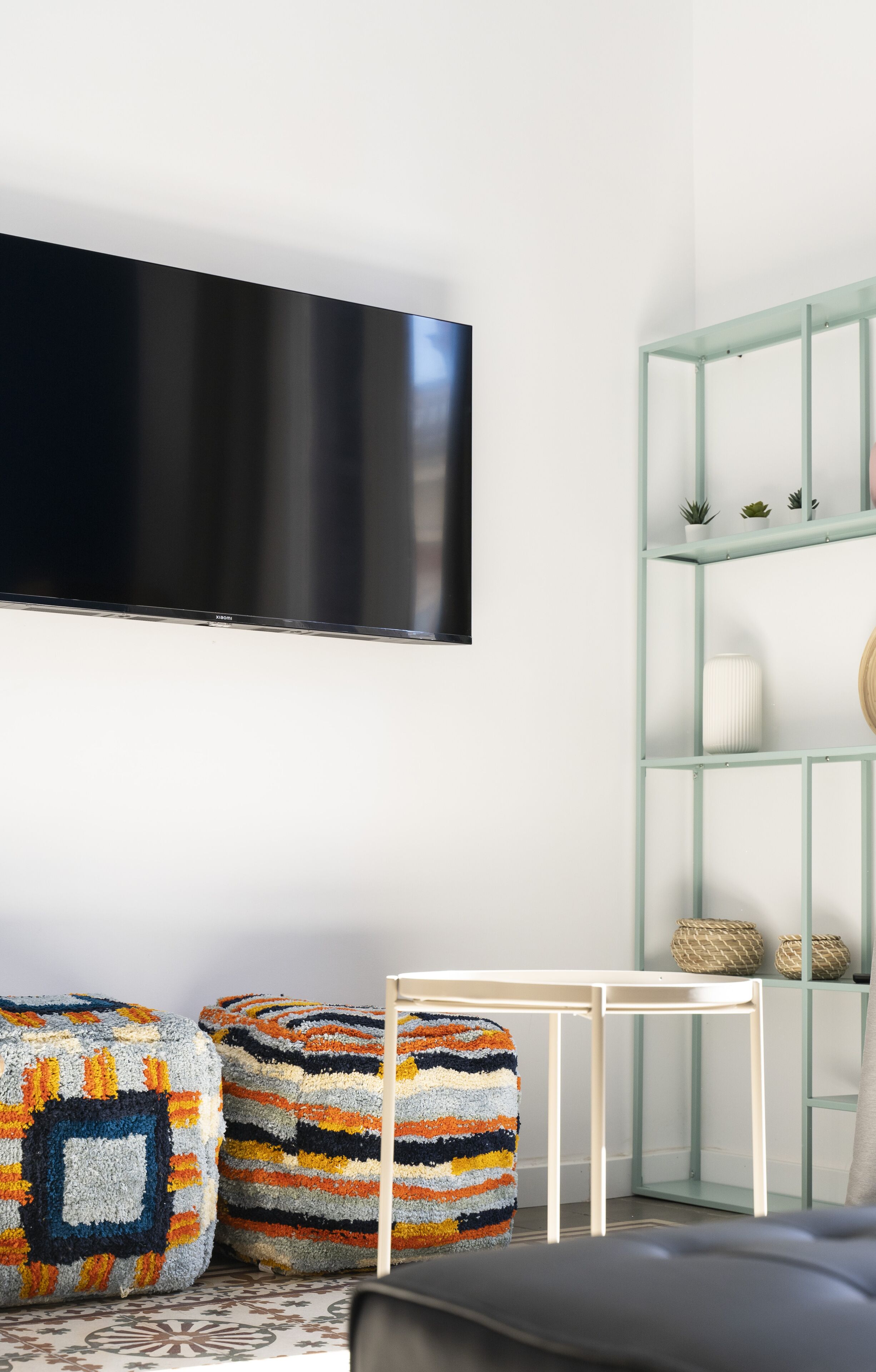 Uma sala de estar moderna com televisão de tela plana na parede, dois pufes coloridos, uma mesa de centro branca e um móvel de estante verde claro com objetos decorativos.