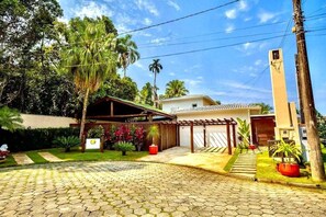 Um imóvel residencial com calçada pavimentada, varanda coberta e jardim com diversas plantas e uma palmeira.
