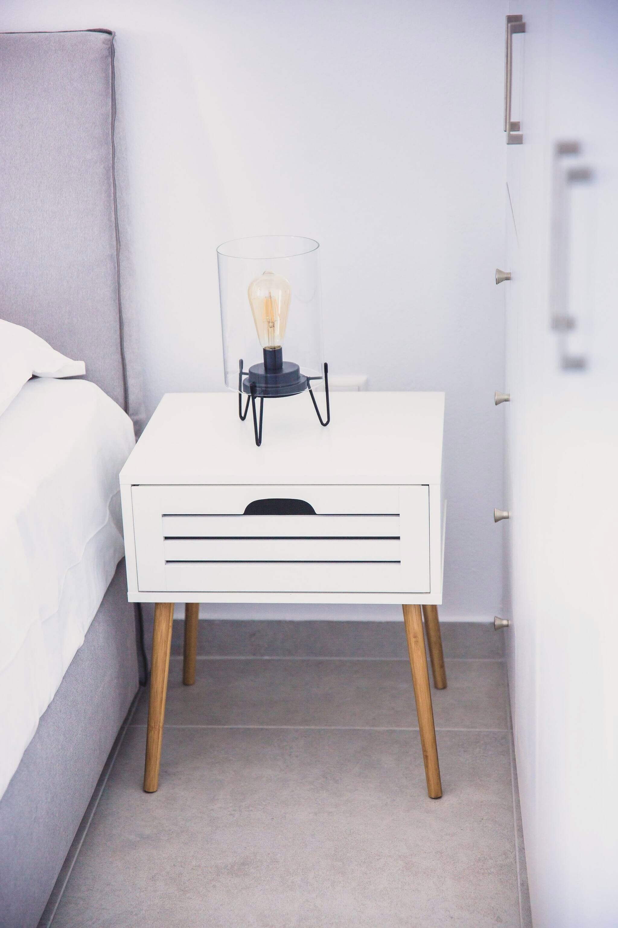 Uma mesa de cabeceira branca com uma luminária de vidro transparente, uma cama com roupa de cama branca e uma cabeceira cinza.