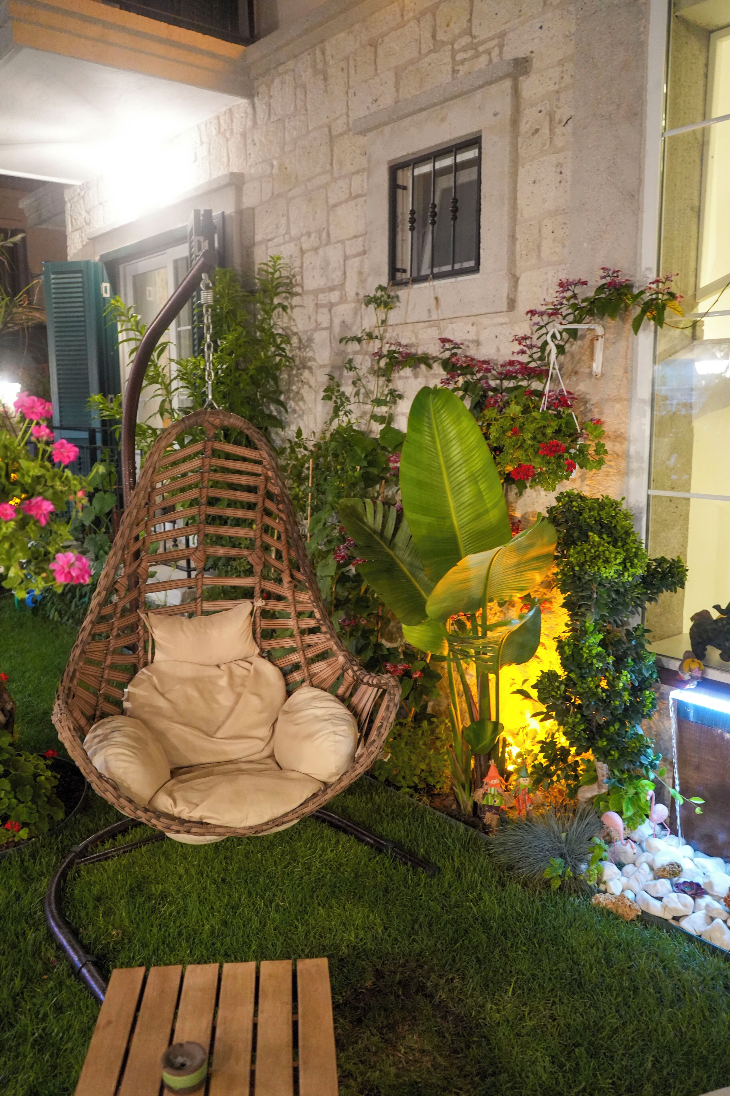 Uma cadeira suspensa num jardim com muro de pedra, plantas exuberantes e uma pequena mesa.