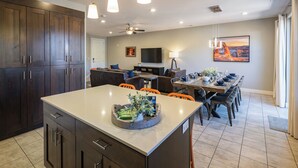 Cozinha moderna com armários de madeira escura, uma ilha central e uma área de refeições com uma mesa comprida e cadeiras laranjas.