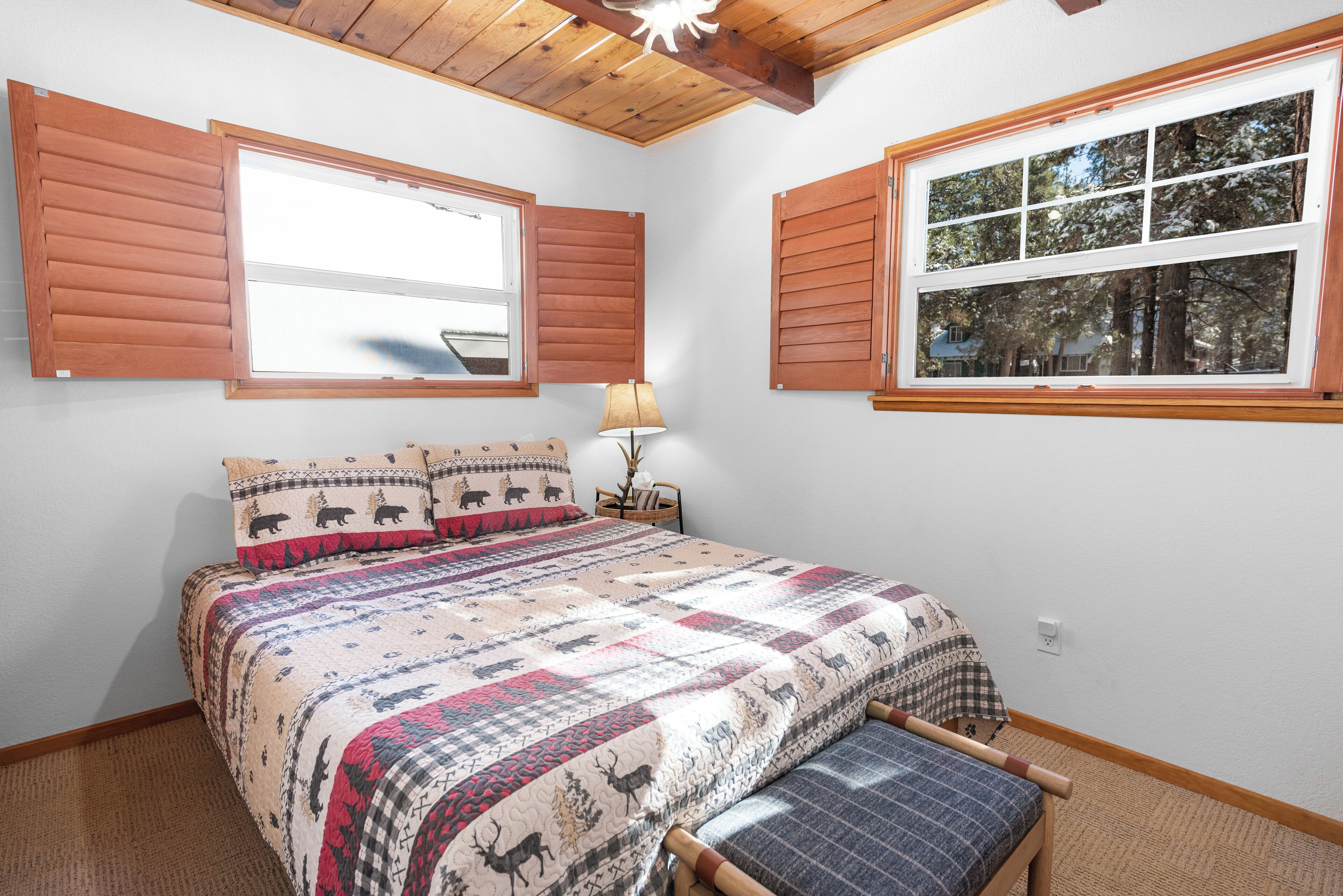Quarto com uma cama grande, persianas de madeira nas janelas, colcha com estampas de animais, abajur e um banco no pé da cama.