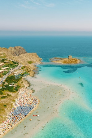 Vista aérea de uma área costeira com praia, uma pequena ilha com uma torre e uma estrada que leva à praia.