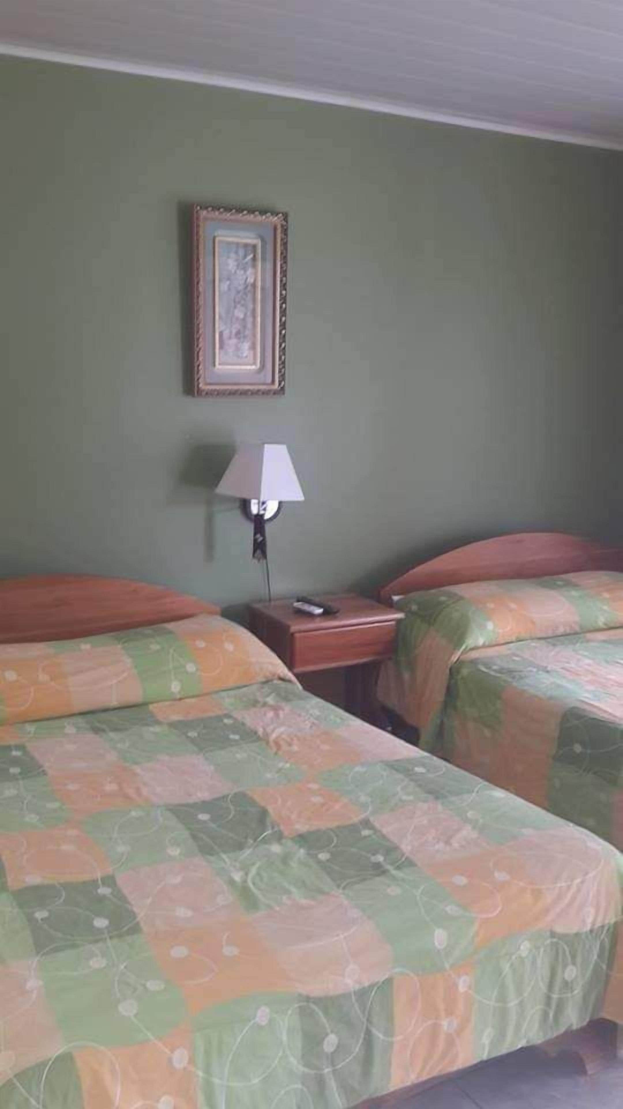 Quarto de hotel com duas camas, uma mesa de cabeceira com abajur, e um quadro na parede.