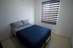 Uma cama de solteiro com colcha azul, um travesseiro e uma janela com persianas horizontais.