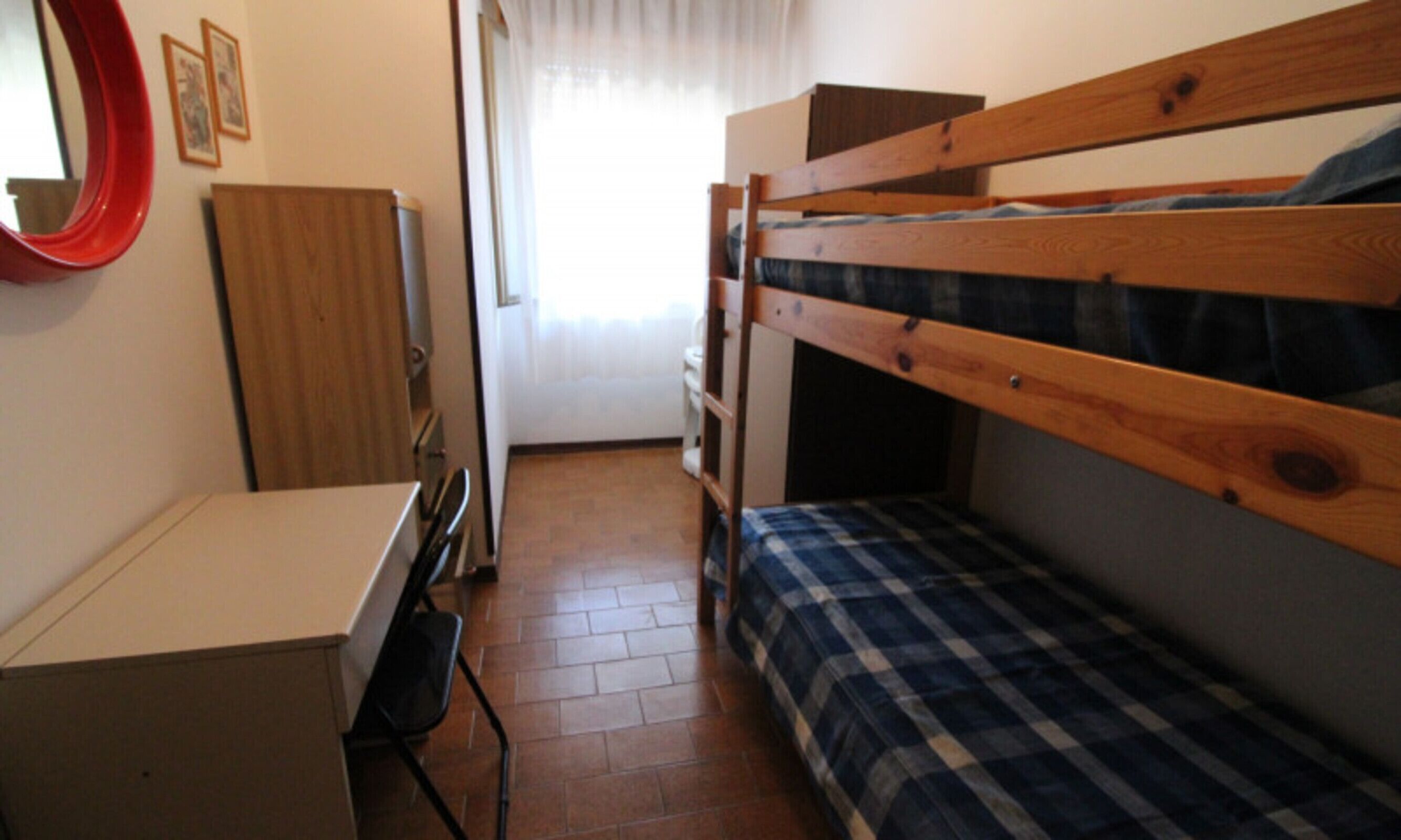 Um dormitório com beliches, uma mesa, uma cadeira, um espelho e uma janela com cortinas.