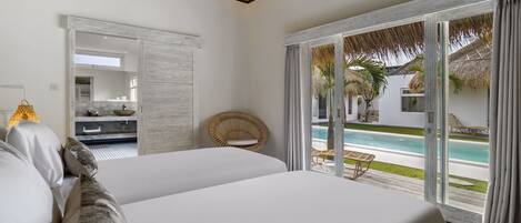 Quarto com uma cama grande, banheiro com espelho e vista para uma piscina e área externa com assentos.