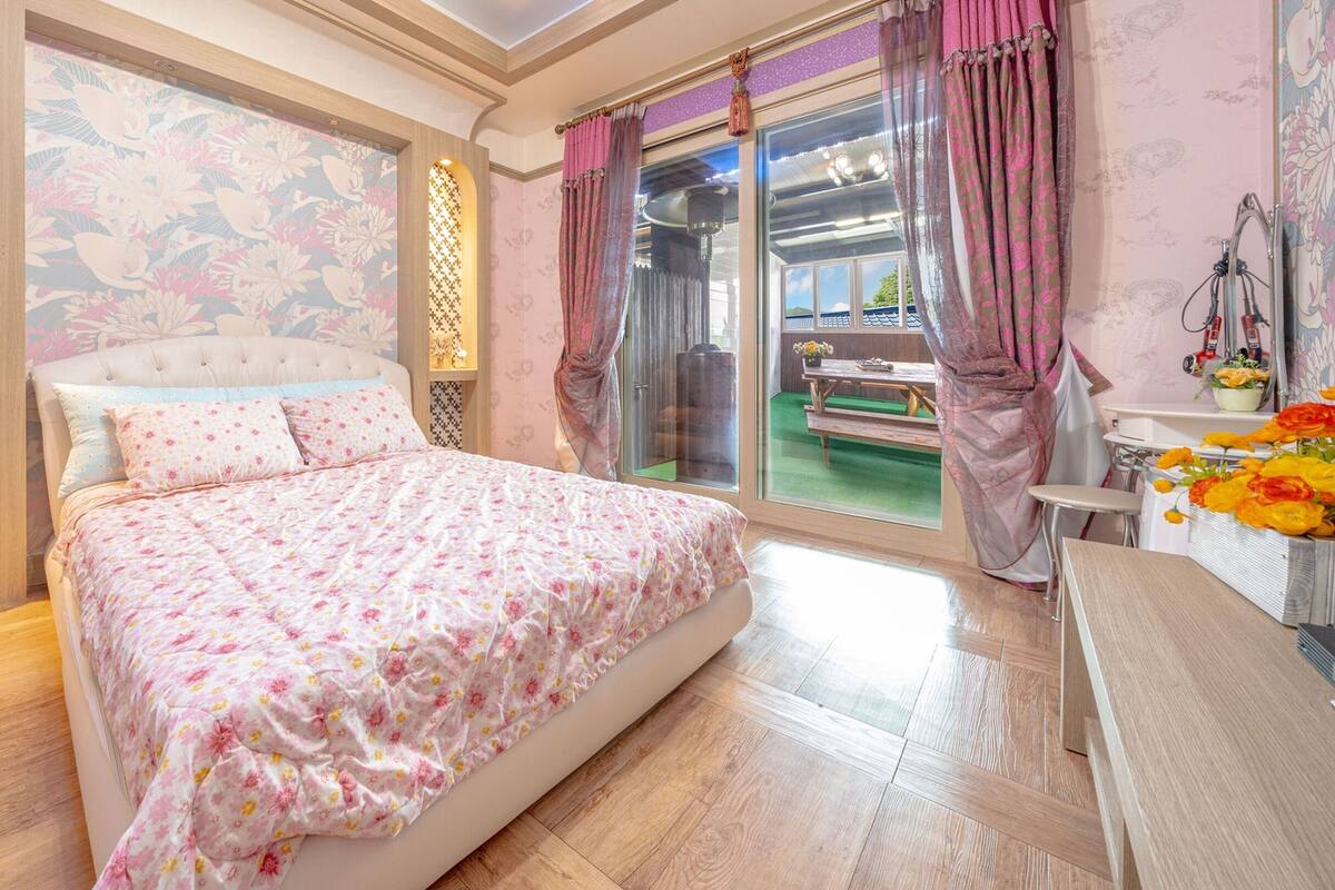 Quarto com cama grande, porta de correr de vidro para uma varanda e uma pequena escrivaninha com um vaso de flores.