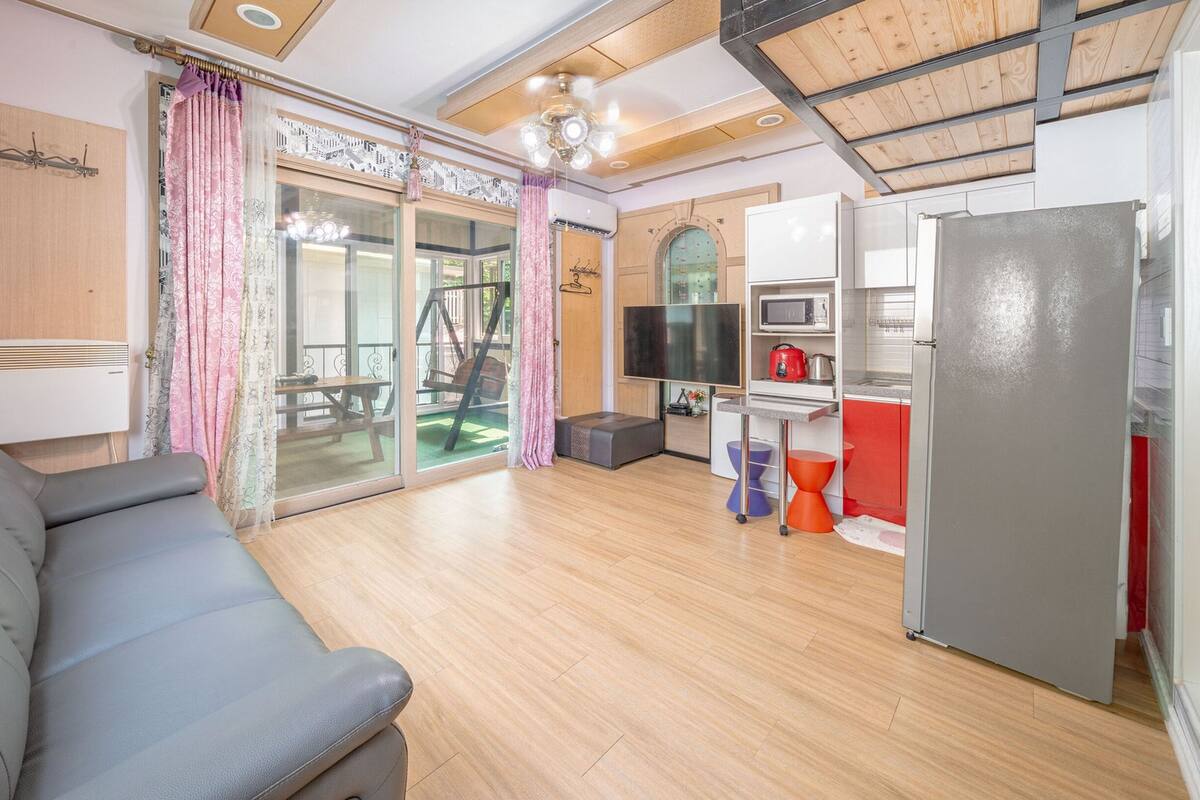 Uma sala de estar moderna com um sofá cinzento, teto de madeira, televisão de ecrã plano, zona de cozinha com frigorífico e uma porta de vidro deslizante que dá para uma área exterior.