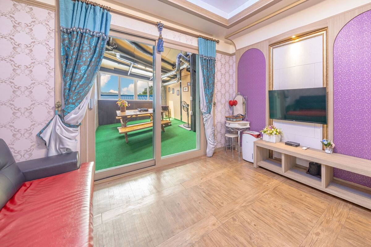 Quarto com piso de madeira, um banco, uma TV e uma janela com cortinas.