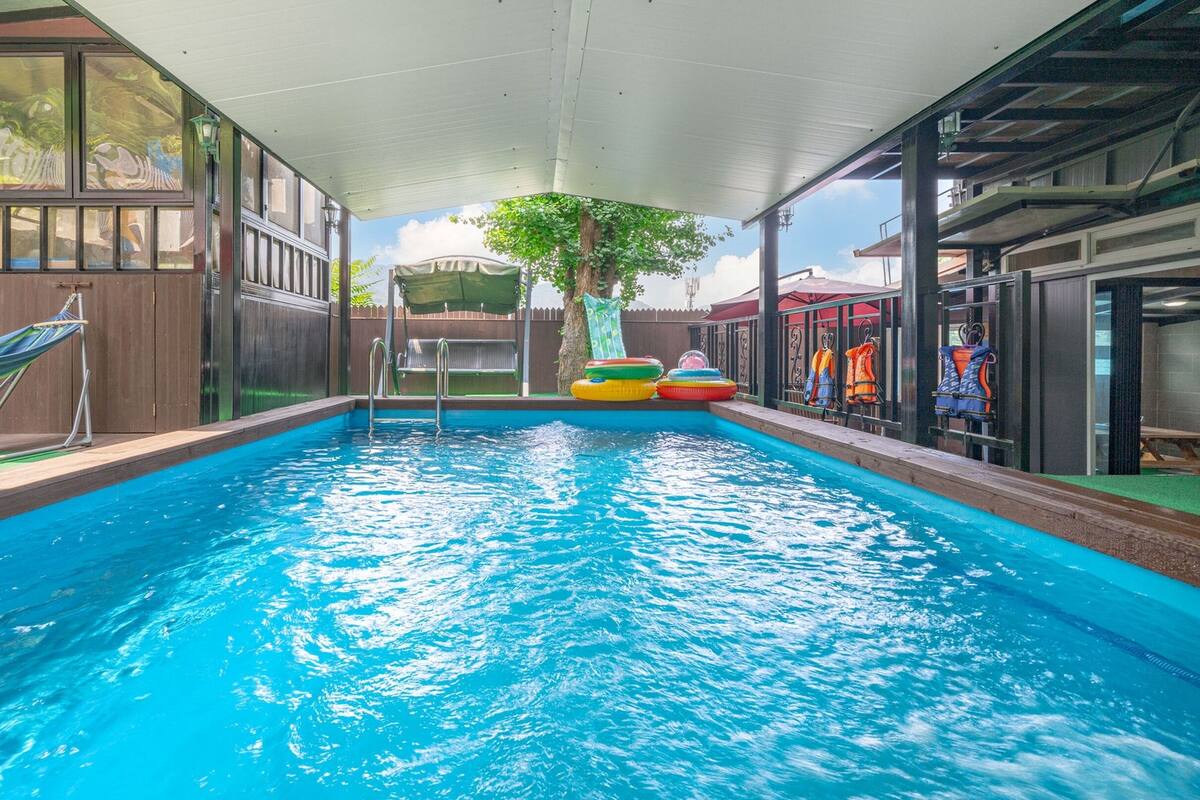 Piscina interior com água azul cristalina, cercada por paredes de madeira e grandes janelas. Há boias coloridas e uma rede próxima à piscina.