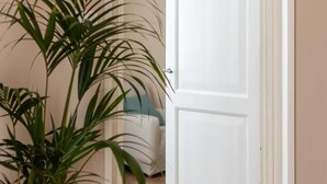 Um corredor com uma porta branca, um vaso de planta e um vislumbre de um quarto com uma cama e uma cadeira azul.