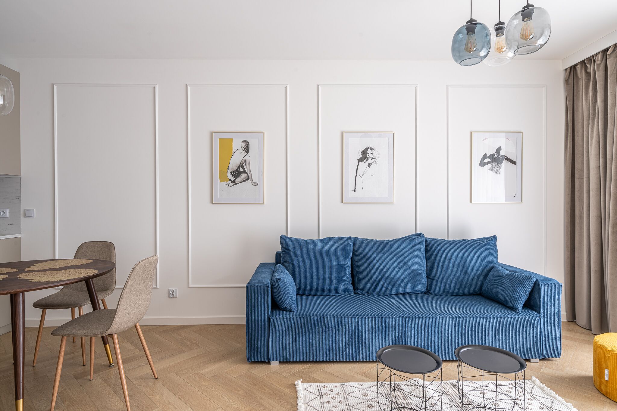 Uma sala de estar moderna com um sofá azul, uma mesa de jantar com cadeiras e três obras de arte emolduradas na parede.