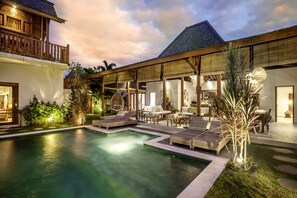 Uma área de piscina com espreguiçadeiras e espaço para refeições, cercada por vegetação exuberante e um edifício grande com varanda.