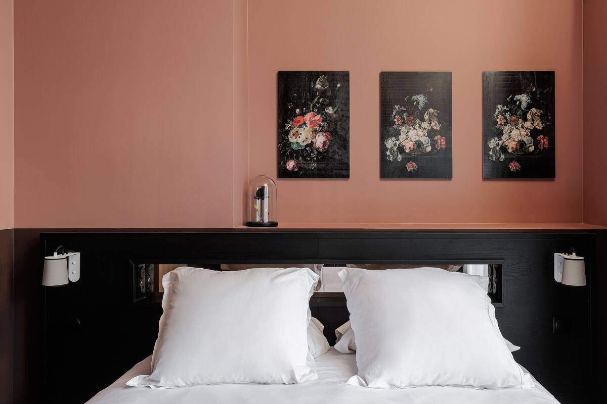Uma cama bem arrumada com lençóis brancos e duas almofadas, cabeceira de madeira escura e três quadros florais na parede.