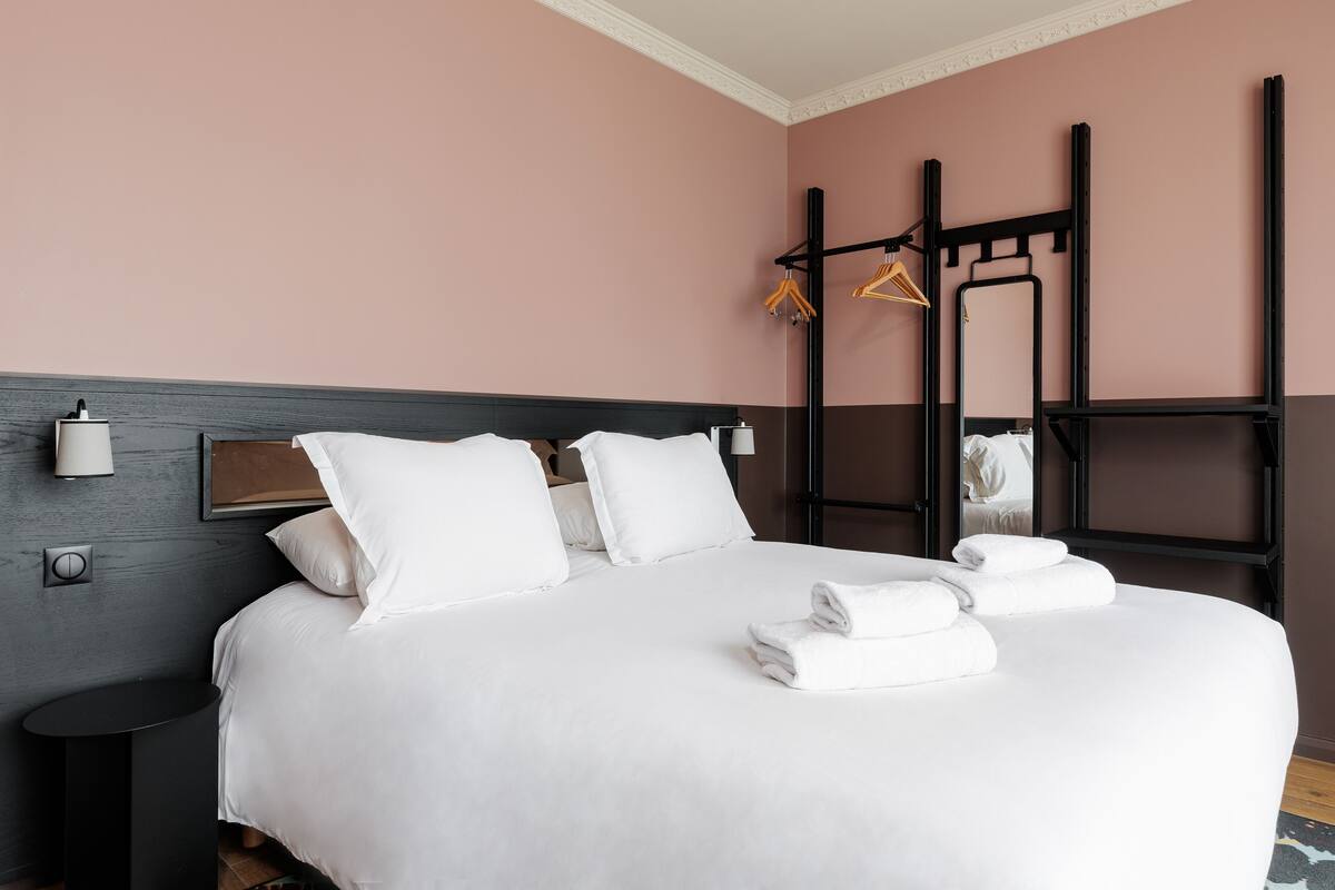 Cama bem arrumada com lençóis e toalhas brancos, uma mesa de cabeceira com abajur e um cabideiro fixado na parede com cabides.