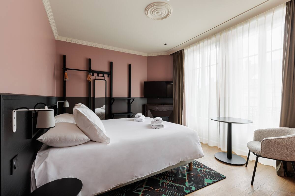 Quarto de hotel com cama, televisão, cadeira, mesa pequena e janela com cortinas.