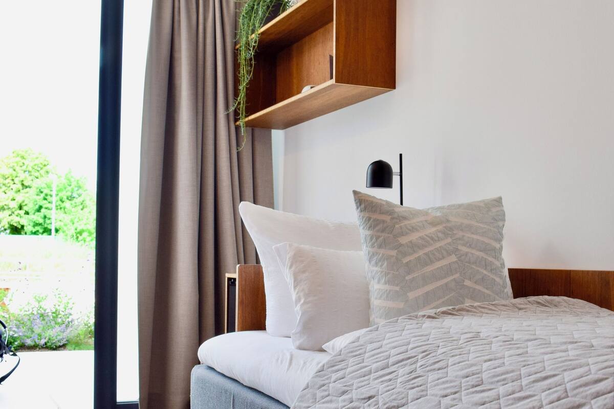 Quarto moderno com cama, uma prateleira de madeira acima dela e vista para o verde lá fora.