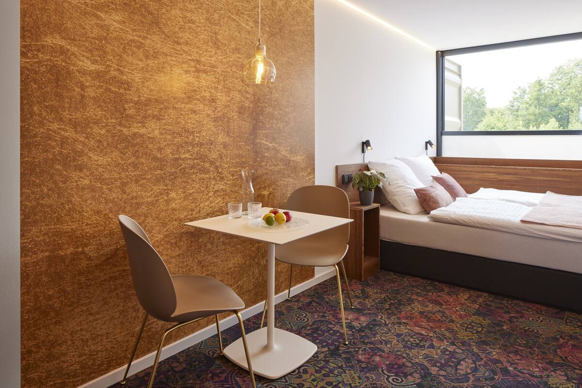 Quarto de hotel moderno com parede de destaque em madeira, área de jantar com mesa e cadeiras, cama com vista para árvores e tapete estampado.