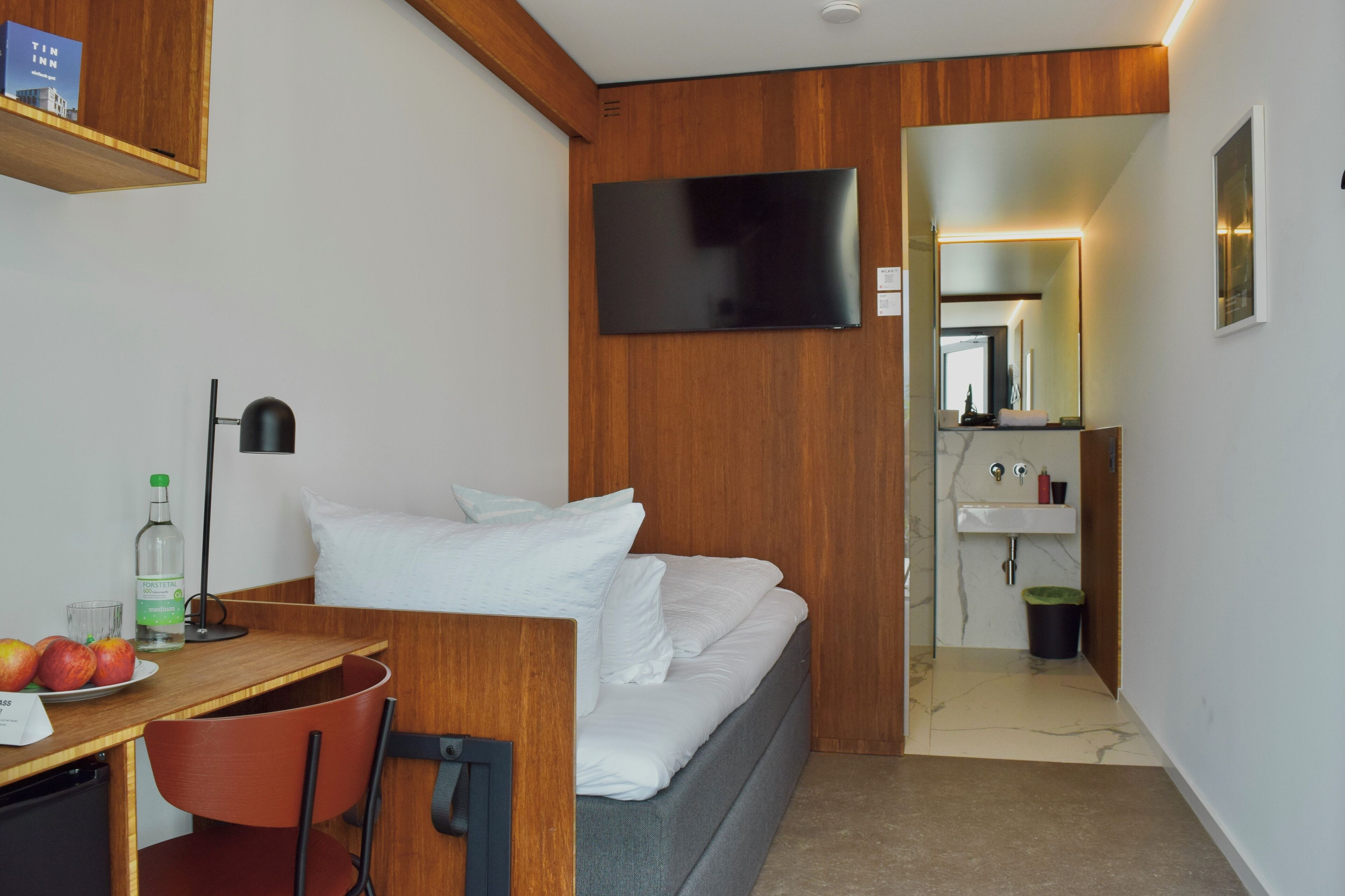 Quarto de hotel com cama, escrivaninha com cadeira, televisão na parede e banheiro com pia e espelho.