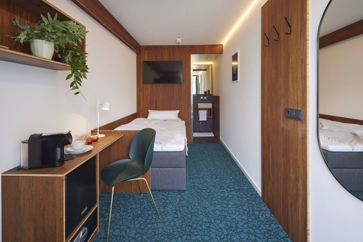 Quarto de hotel compacto com cama, escrivaninha com cadeira, pequeno espaço de cozinha, espelho e planta em vaso.