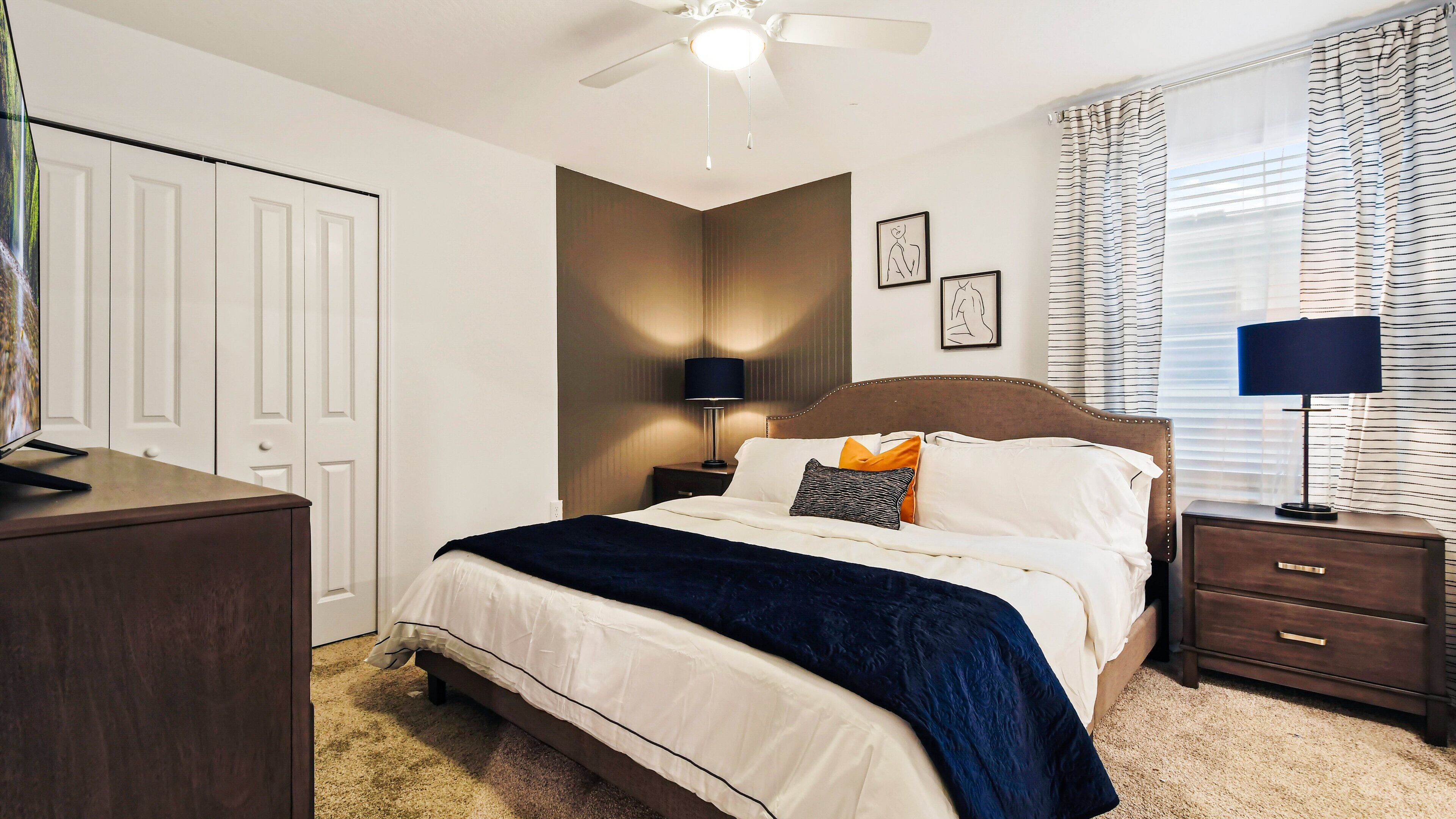 Quarto com cama, mesas de cabeceira, escrivaninha, ventoinha no teto e janela com cortinas.
