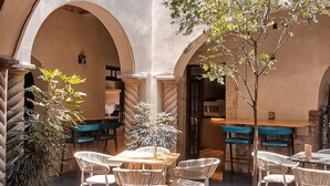 Um café ao ar livre com arcos, mesas e cadeiras, rodeado por vegetação e uma árvore.