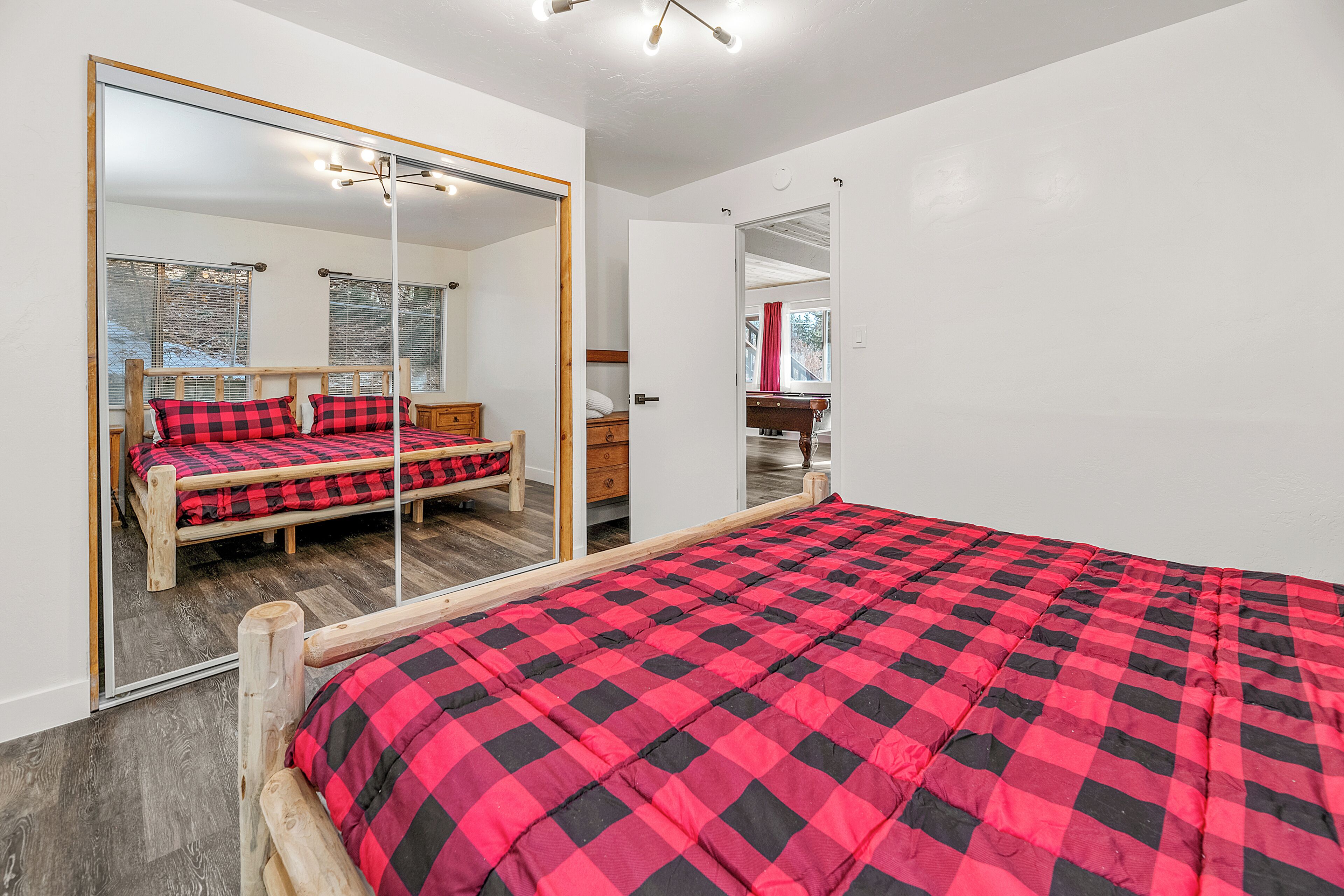 Quarto com um espelho grande, cama com colcha xadrez vermelha e preta, cabeceira de madeira e chão de madeira.