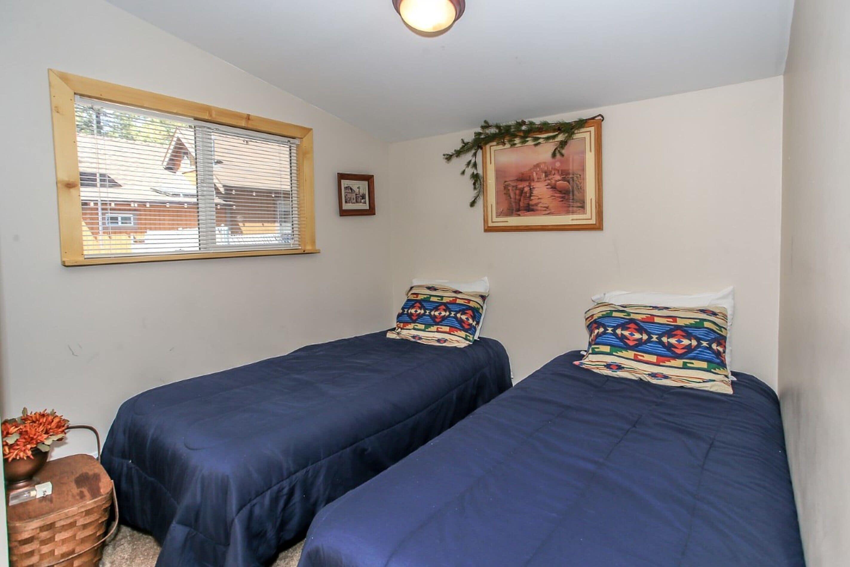 Quarto com duas camas, janela com persianas, um quadro emoldurado e uma pequena planta.
