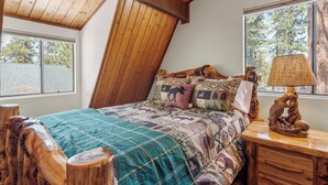 Quarto com teto inclinado, uma janela grande, cama com roupa de cama estampada, mesa de cabeceira de madeira e um abajur.