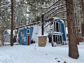 Uma cabana coberta de neve com chaminé de pedra, porta vermelha e uma grande janela.