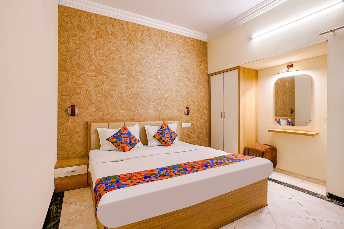Quarto de hotel com uma cama grande, cabeceira de madeira, parede estampada, um banquinho pequeno e um espelho acima da cama.
