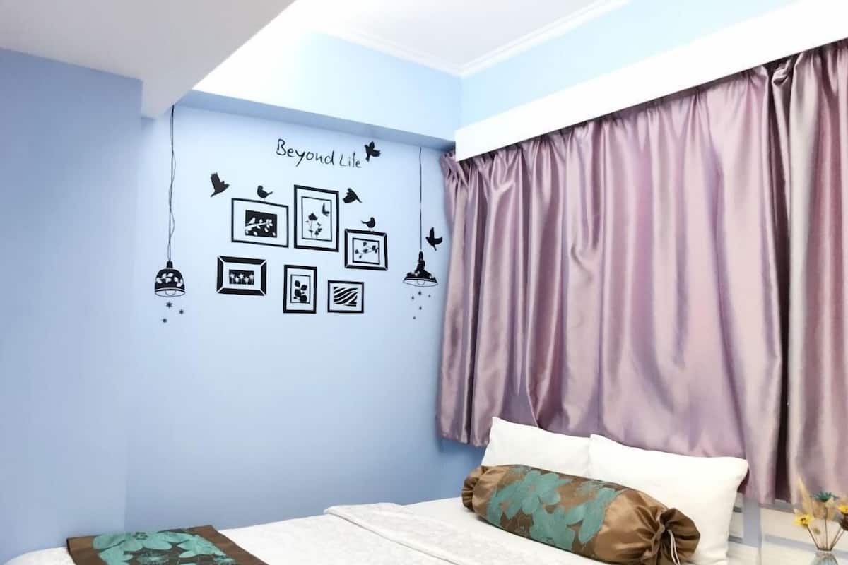Quarto com cama, parede adornada com quadros e janela com cortinas roxas.
