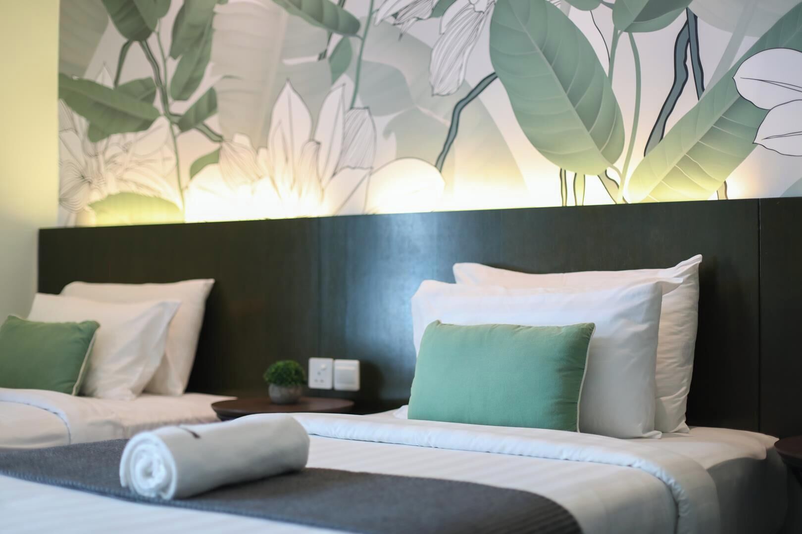 Quarto de hotel com cama, almofadas, uma mesinha com uma planta e uma parede com um mural floral.