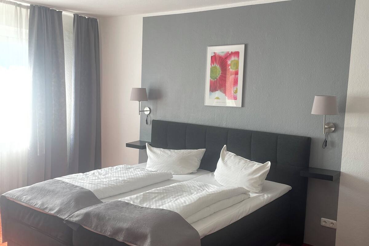 Quarto de hotel com uma cama grande, paredes cinzentas, um quadro emoldurado, duas luminárias de cabeceira e um tapete vermelho.
