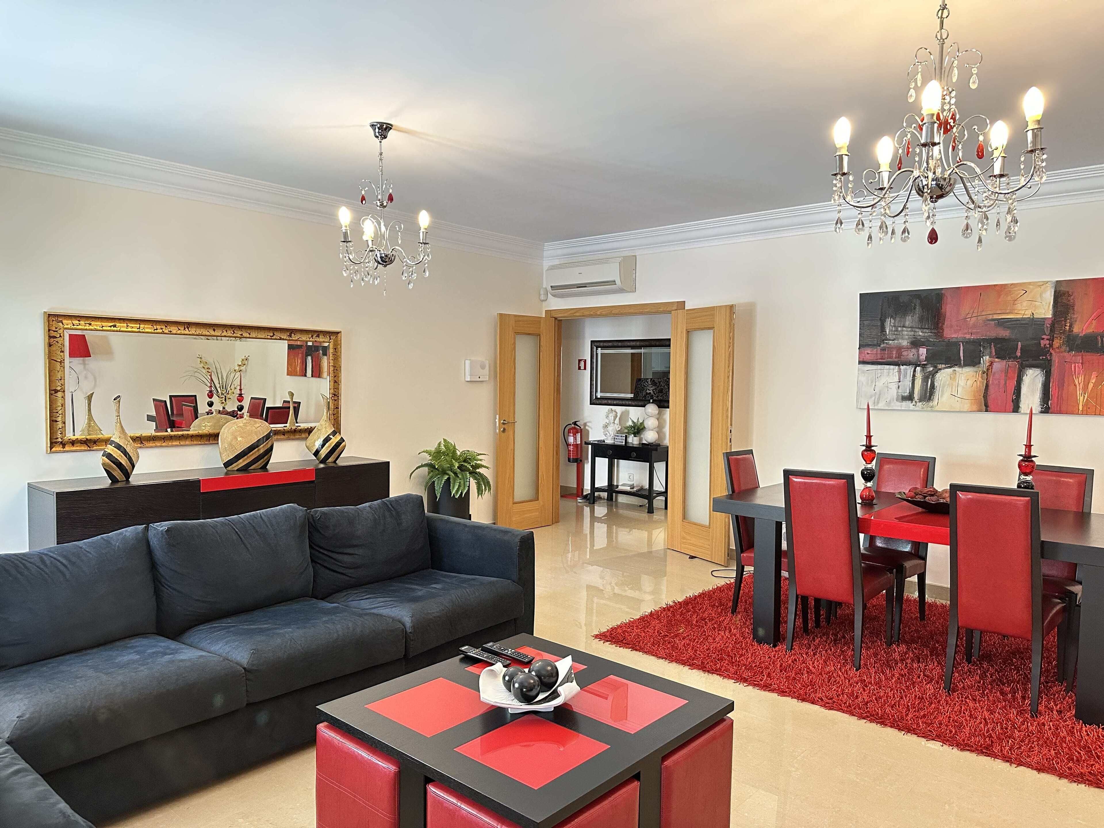 Uma sala moderna com um sofá preto, uma mesa de centro preta e vermelha, e um candelabro. Há um espaço de jantar com cadeiras vermelhas e um tapete vermelho.