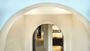 Um corredor com teto curvo e abobadado que leva a um quarto com piso escuro, uma cadeira e quadros nas paredes.