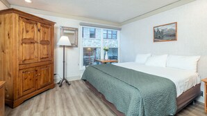 Quarto com uma cama grande, guarda-roupa de madeira, uma escrivaninha com abajur, uma janela com vista e um quadro na parede.