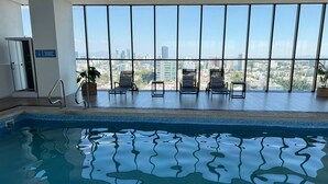 Piscina interior com vista para a cidade, cadeiras de descanso e uma planta em vaso.