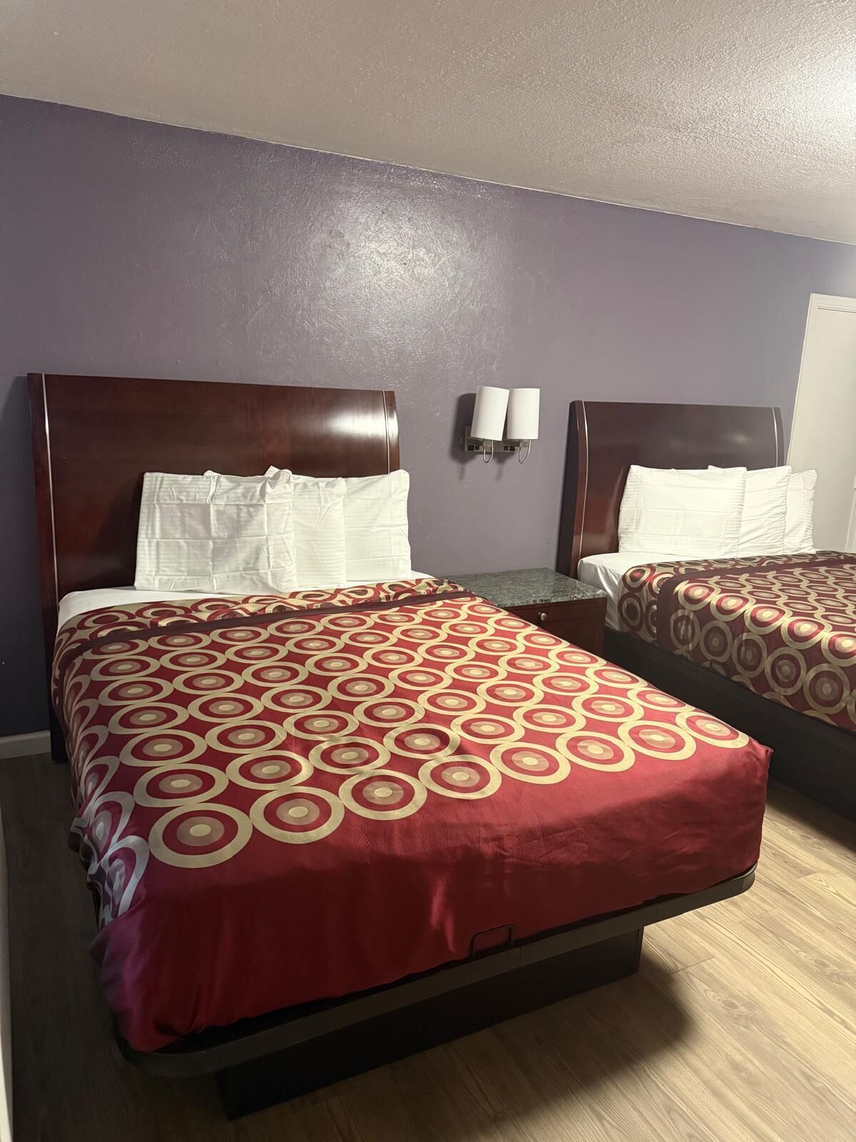 Quarto de hotel com duas camas, cada uma com cabeceira, mesa de cabeceira e luminária fixa na parede.