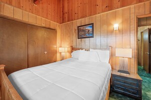 Quarto com painéis de madeira, uma cama, duas luminárias fixadas na parede e um quadro emoldurado sobre a cama.