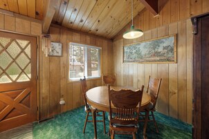 Interior de madeira com uma mesa de jantar e cadeiras, uma janela com persianas, uma porta com vitrais em padrão de losangos e um quadro fixado na parede.