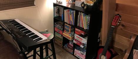 Um quarto com um piano, uma estante cheia de jogos de tabuleiro, uma guitarra e um ursinho de pelúcia.