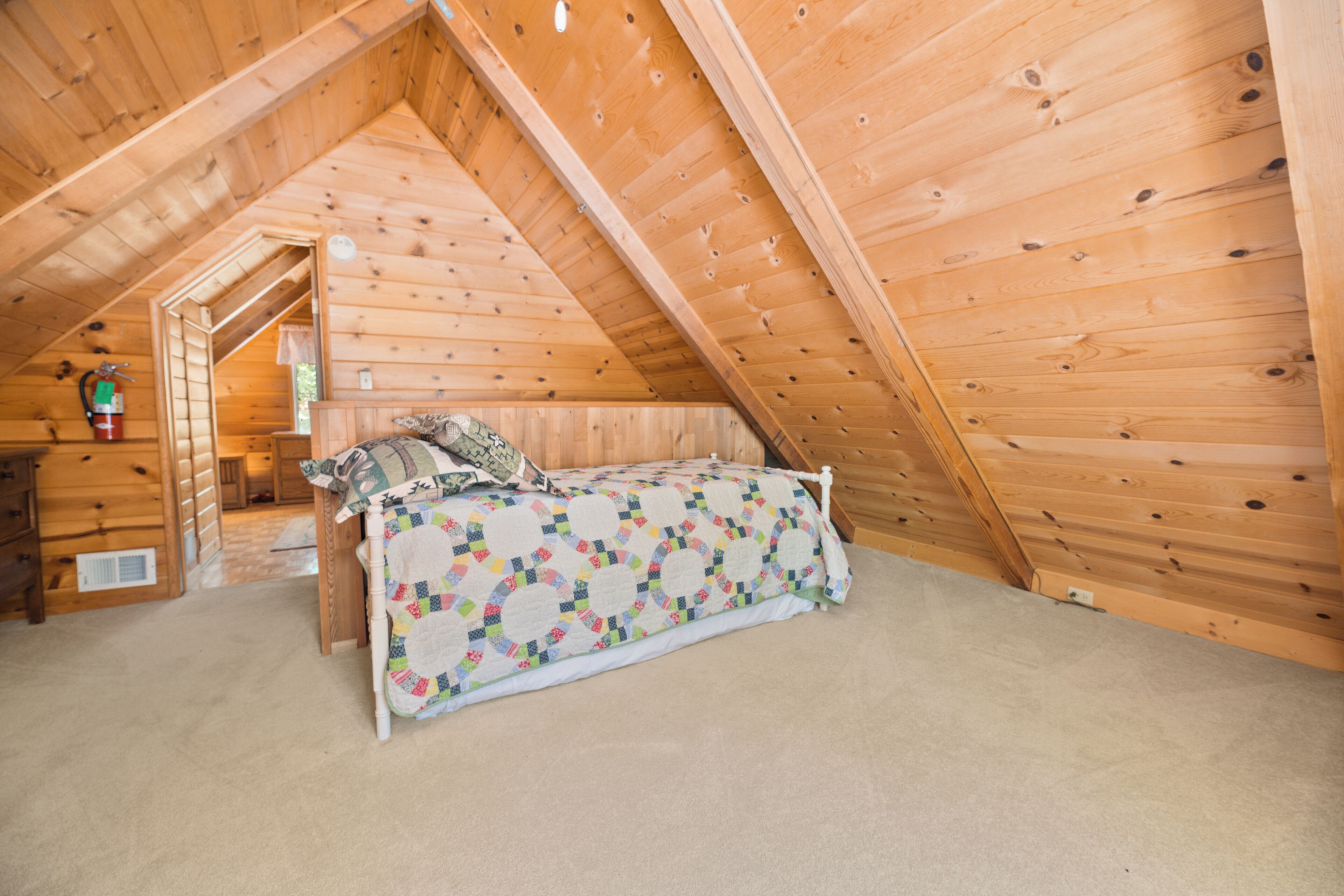 Um quarto de sótão aconchegante, com teto inclinado de madeira, uma cama com colcha colorida e uma pequena cômoda de madeira.