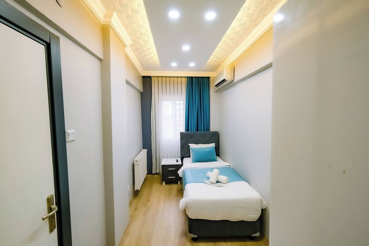 Quarto de hotel estreito com cama, mesa de cabeceira e um banquinho.