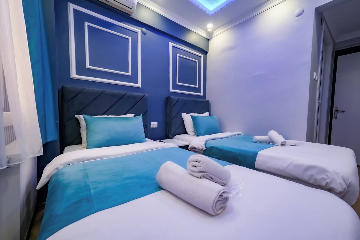 Quarto de hotel com duas camas, cada uma com colcha azul e almofadas brancas. Toalhas dobradas estão sobre as camas. O quarto tem um design moderno, com paredes azuis e uma luminária no teto.
