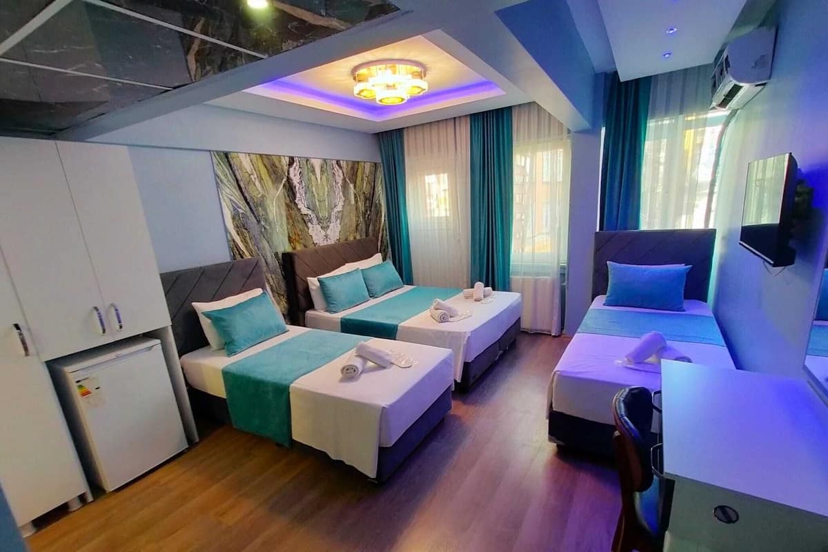 Quarto de hotel com duas camas, uma escrivaninha, uma pequena cozinha e um teto com luminária.
