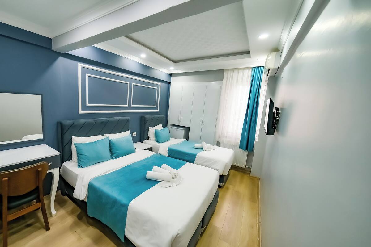 Quarto de hotel com duas camas, uma escrivaninha, uma cadeira, uma televisão e um guarda-roupa.