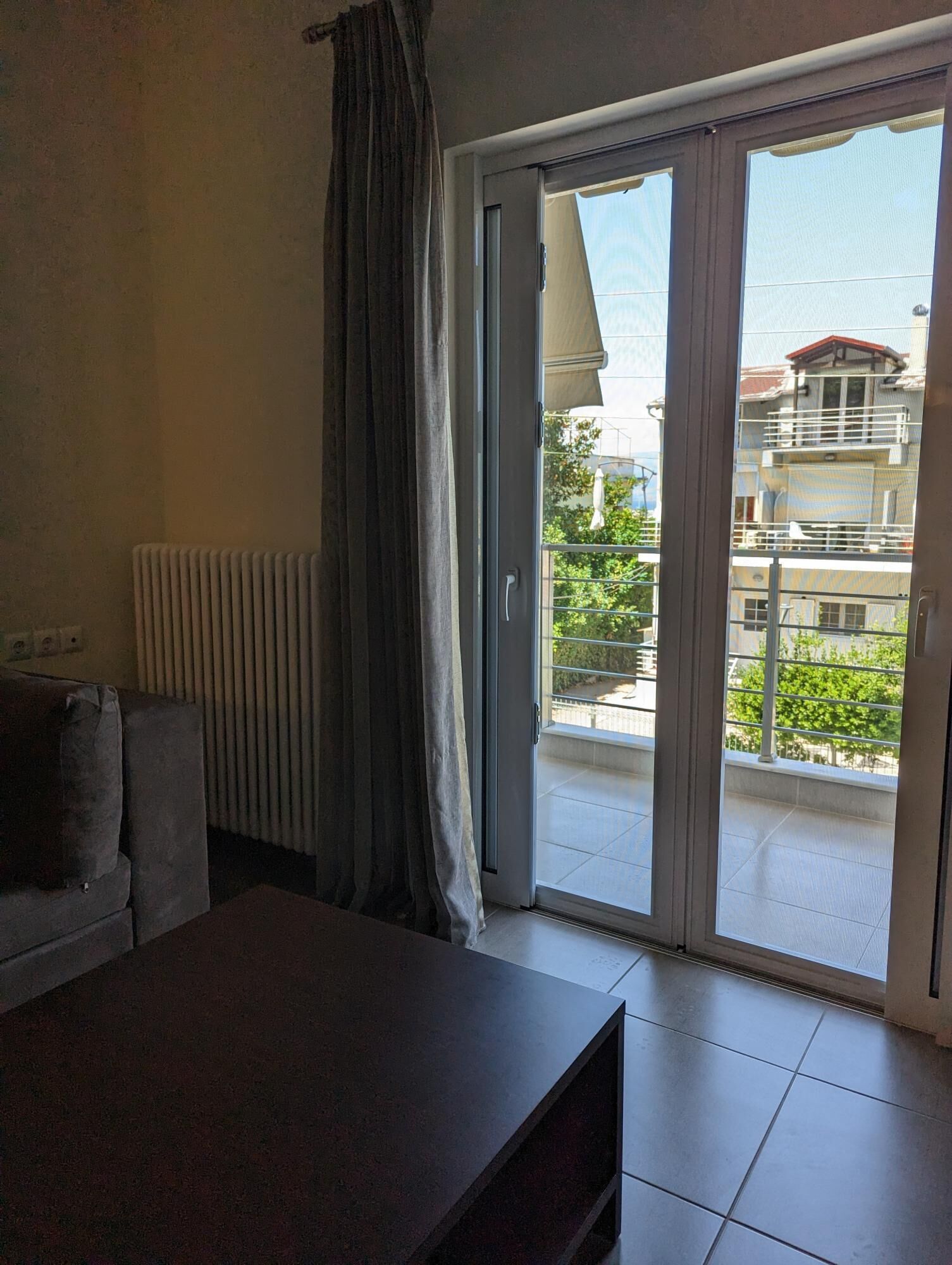 Quarto com sofá escuro, radiador e porta de correr de vidro que dá para uma varanda com vista para edifícios e vegetação.