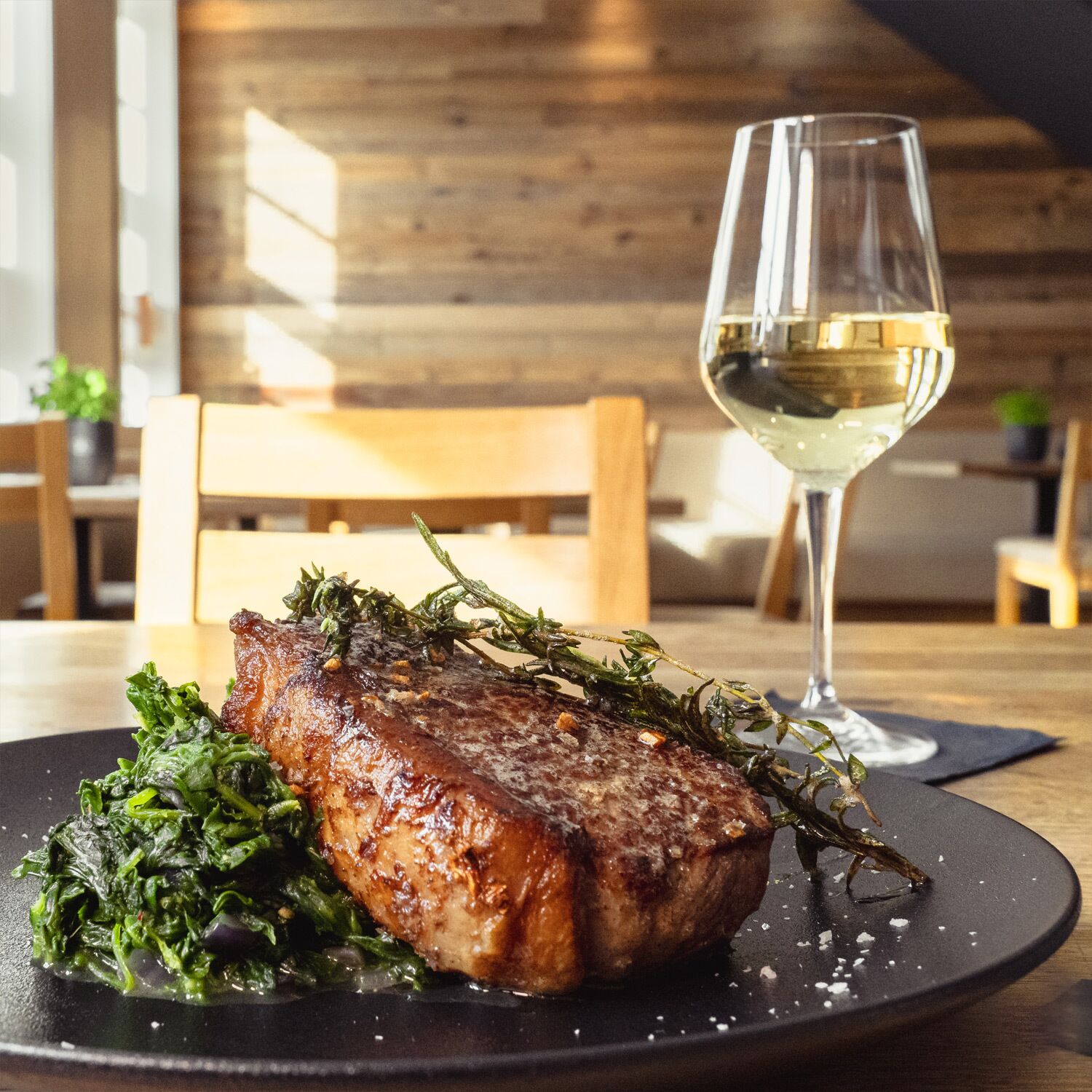 Prato com bife grelhado, vegetais verdes e um copo de vinho branco sobre uma mesa em ambiente de madeira.